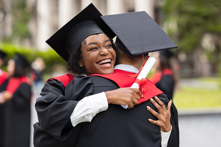 Black female graduate in a cap and gown smiling and hugging another ...